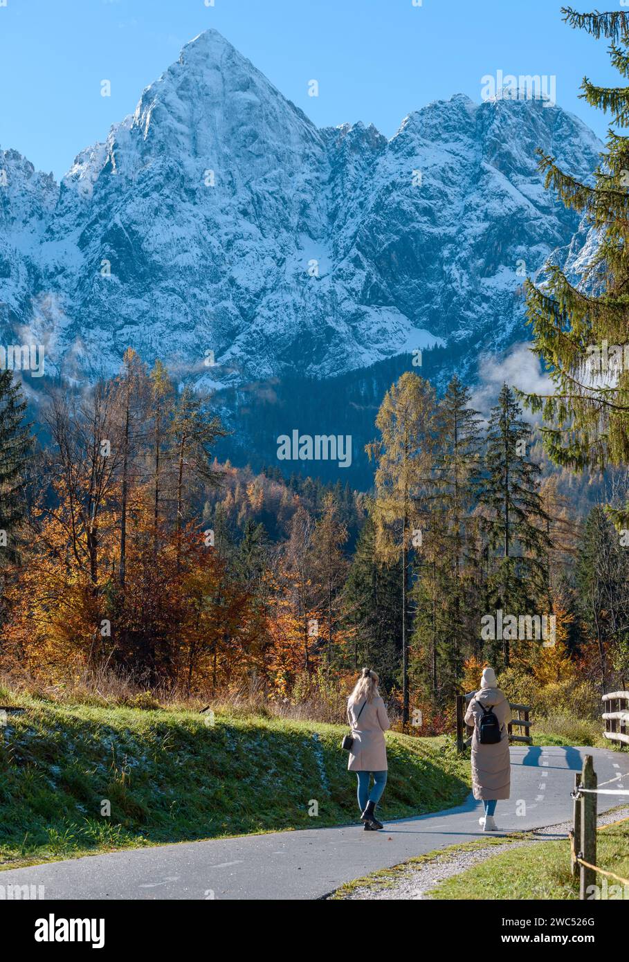 Two women walking on a trail under snowy mountain peaks on autumn day ...