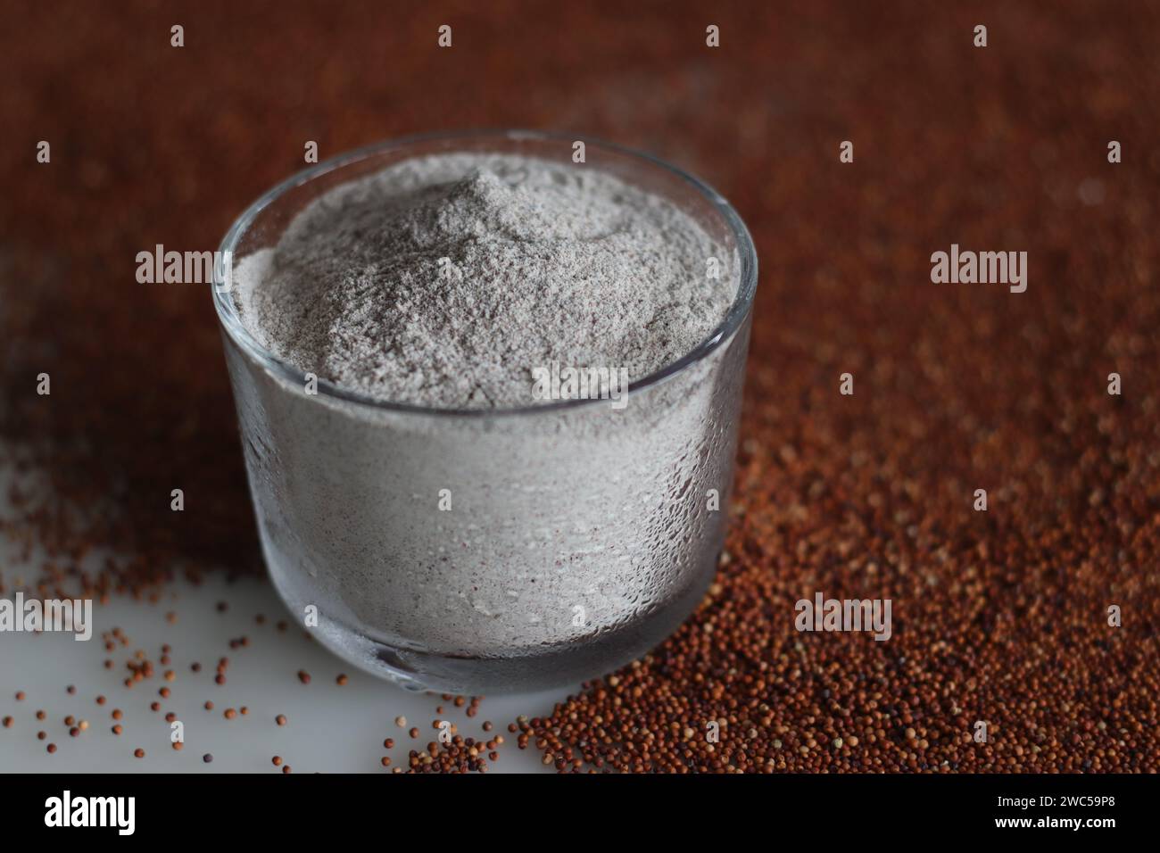 Ragi flour in a glass bowl along with a scattered spread of ragi grains ...