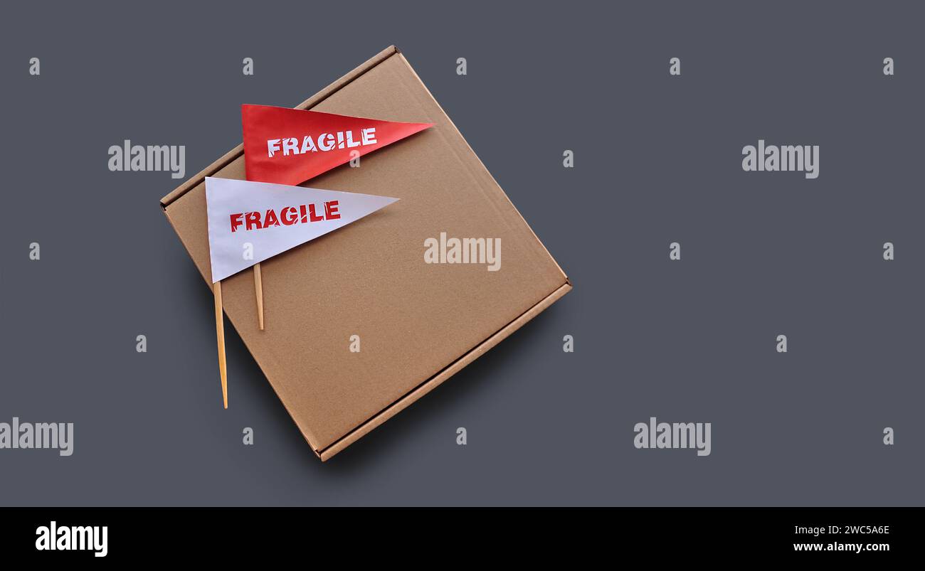 Small paper flags with the warning 'Fragile' as a label and empty cardboard box on monochrome ...