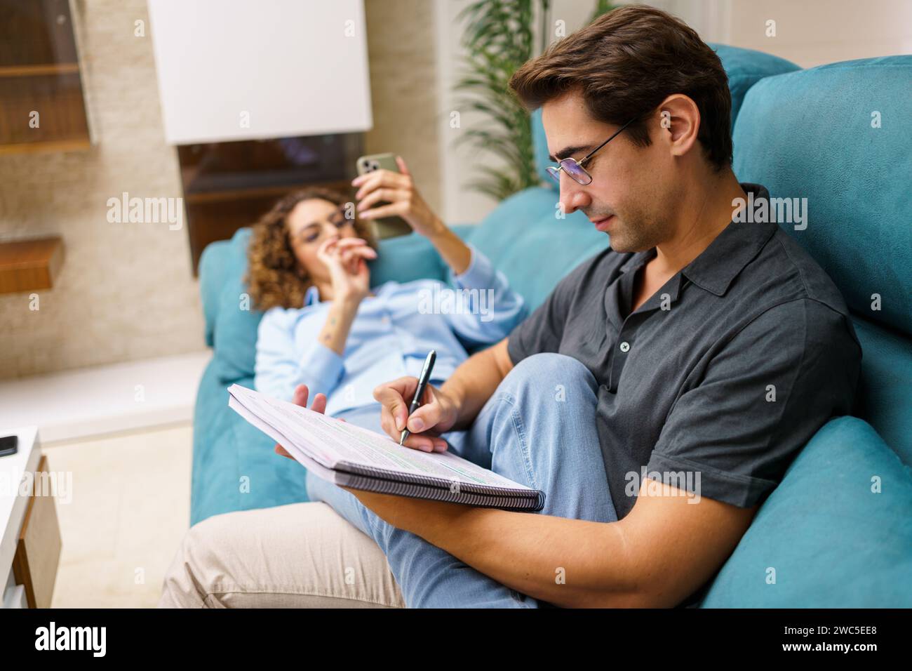 Focused young husband taking notes in notebook while sitting with wife ...