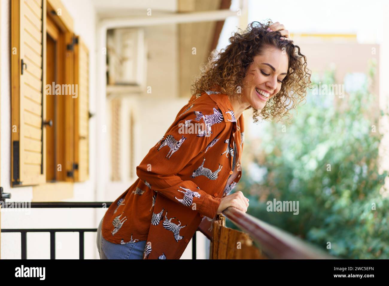 Smiling young woman looking down while standing on balcony of house ...
