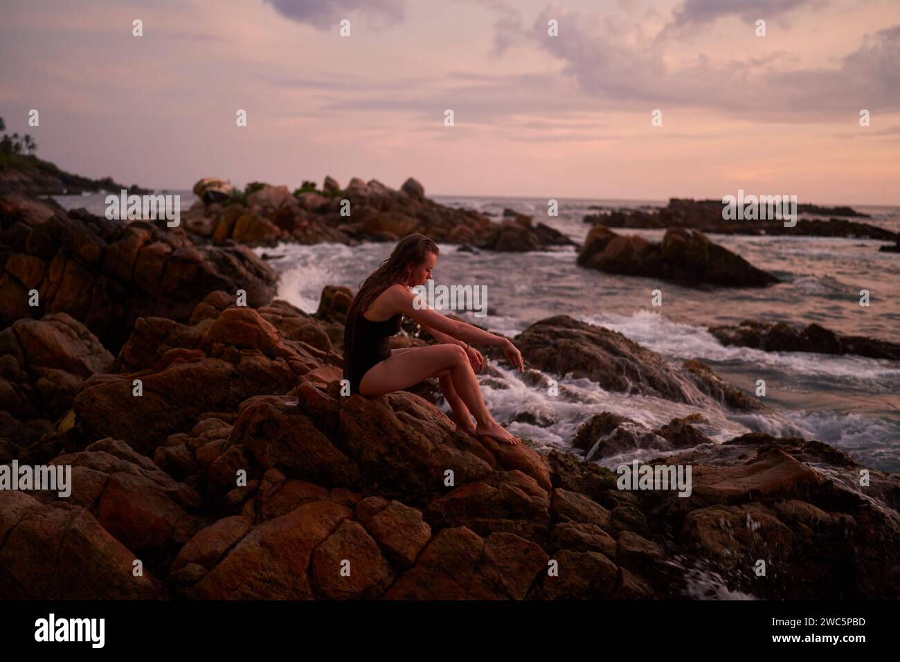 Woman relaxes on seaside rocks at dusk, legs dipped in splashing ocean ...