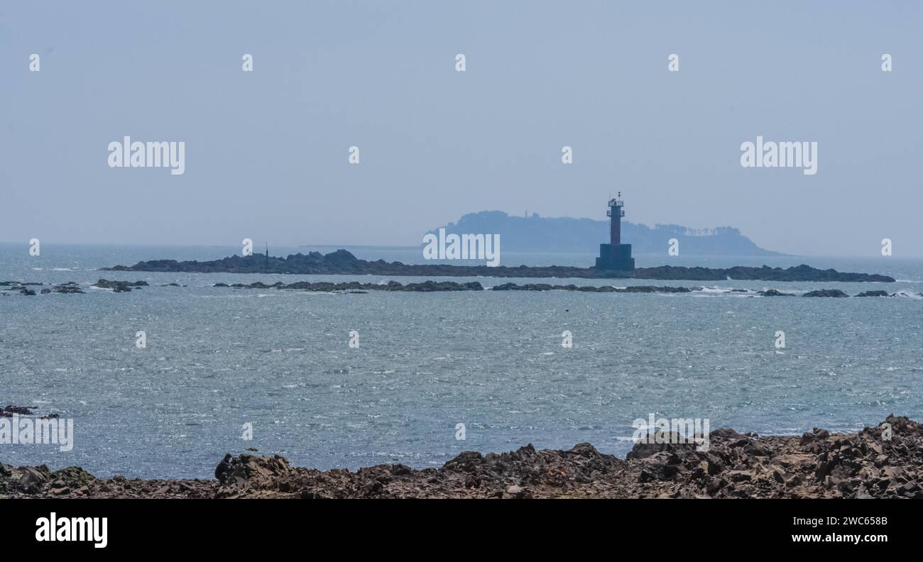 Small lighthouse on rocky outcrop in ocean with a small island obscured ...