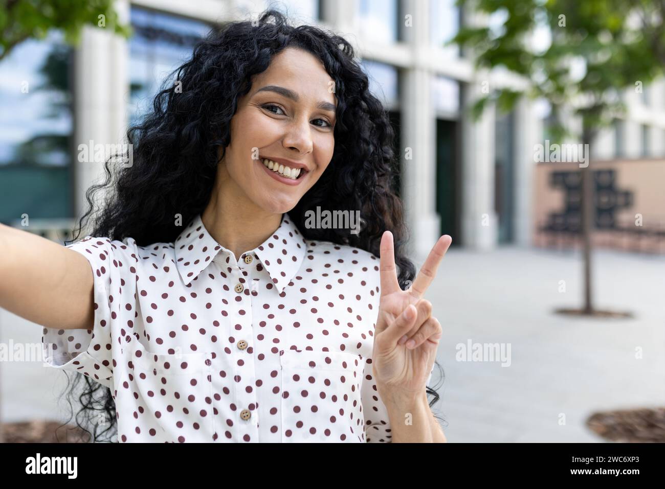 Cheerful young woman with curly hair taking a selfie, flashing a peace ...