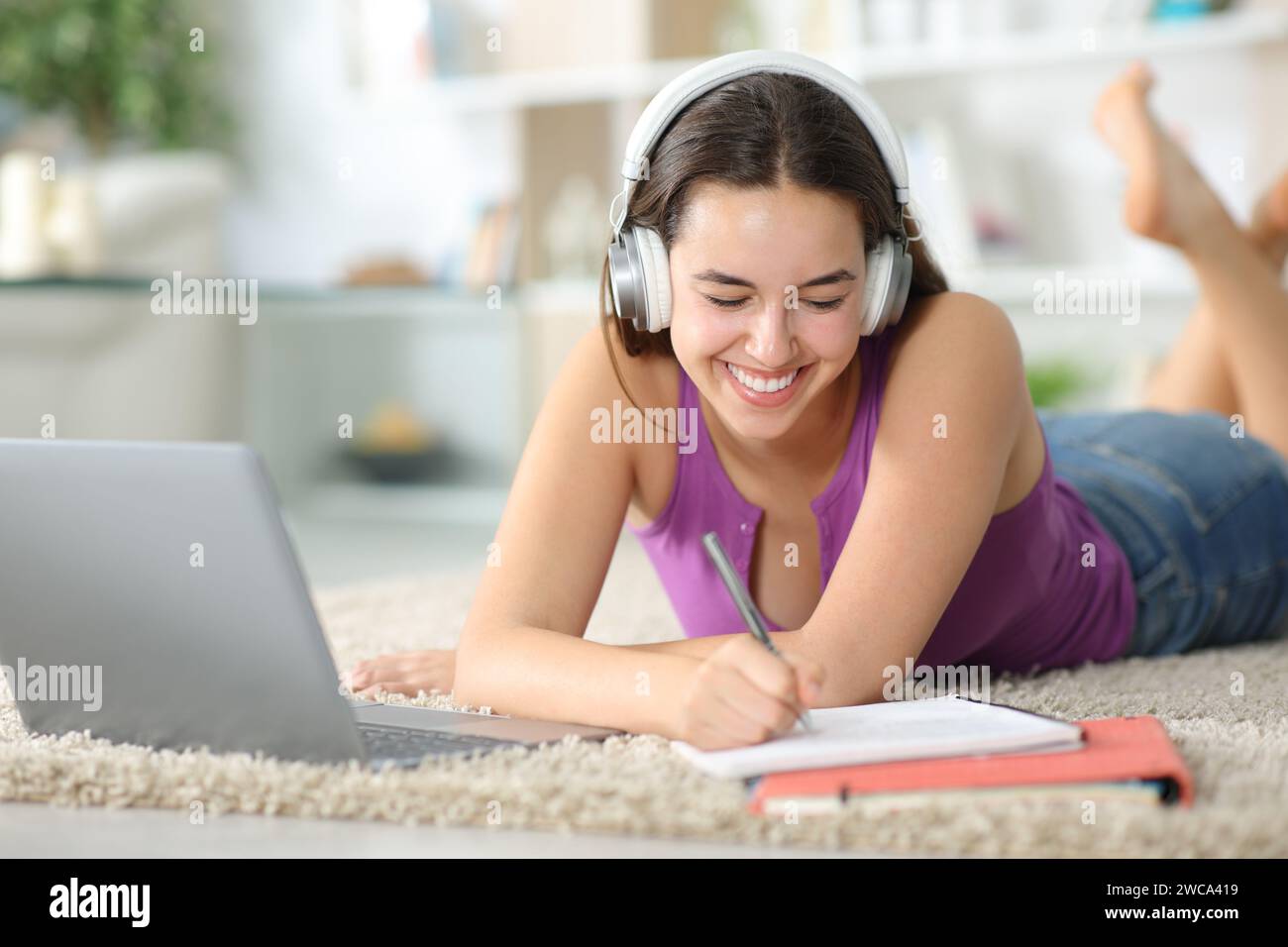 Happy student e-learning and taking notes lying on the floor at home ...