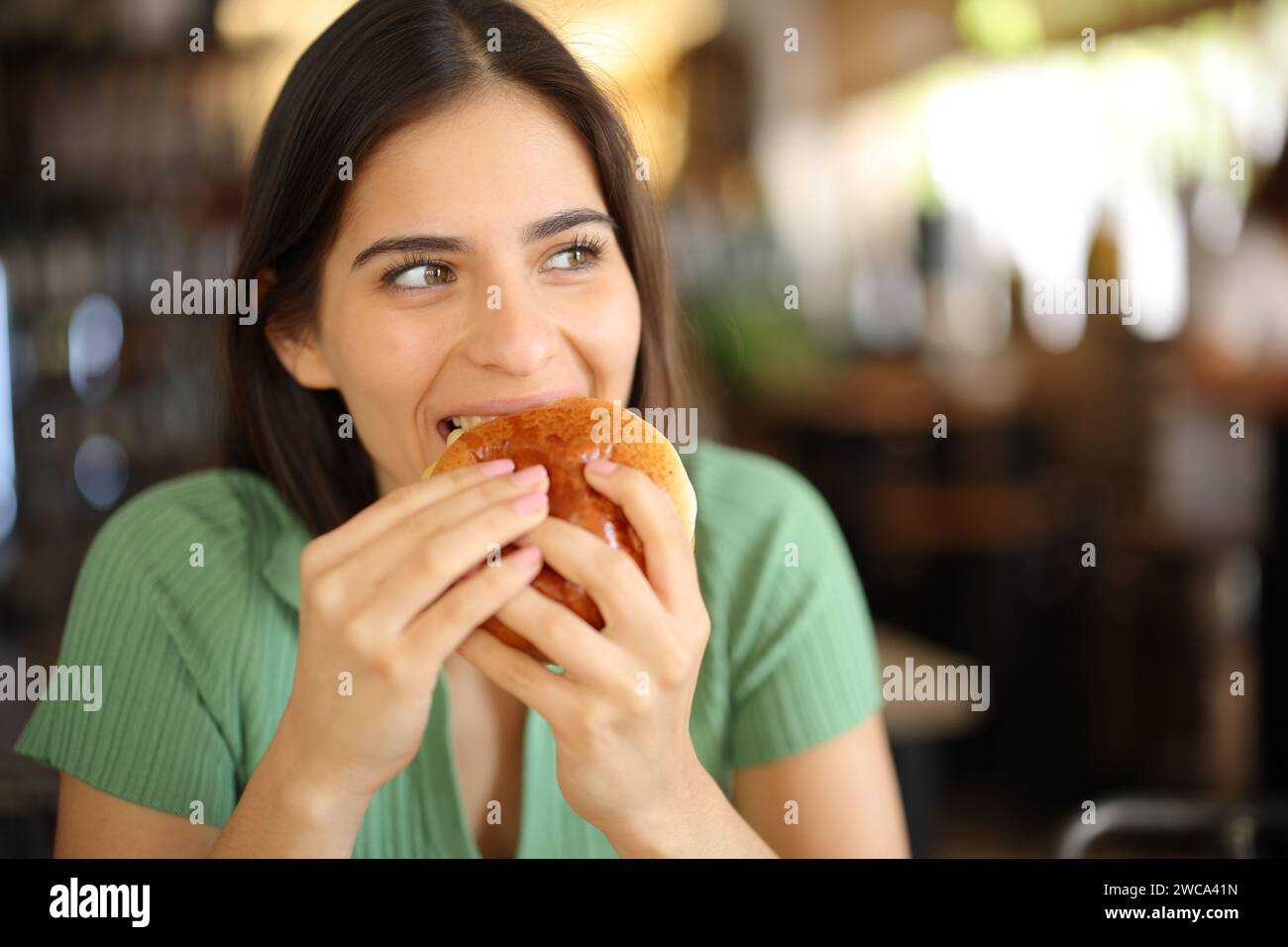 Happy woman eating burger looking at side in a restaurant interior ...