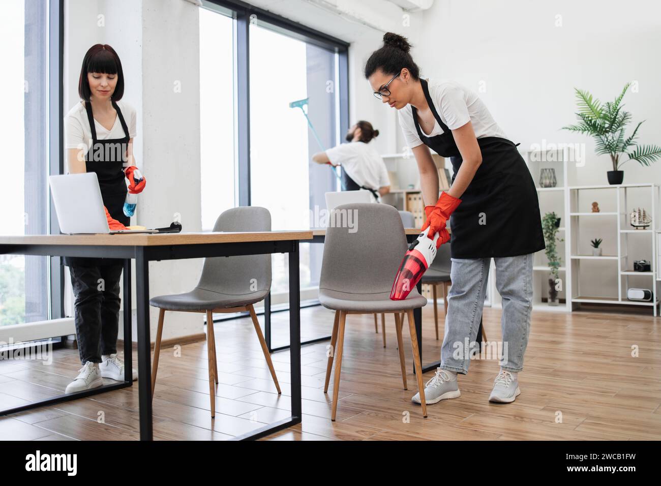 Multiethnic female cleaner vacuuming chair with portable vacuum cleaner ...