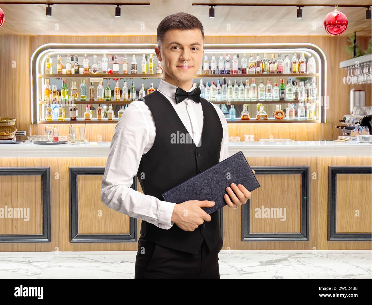 Young server with a bow tie holding a menu in front of a bar with ...