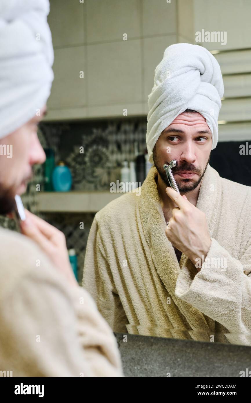 Reflection in mirror of young bearded man in bathrobe using Y-shaped ball roller while massaging ...
