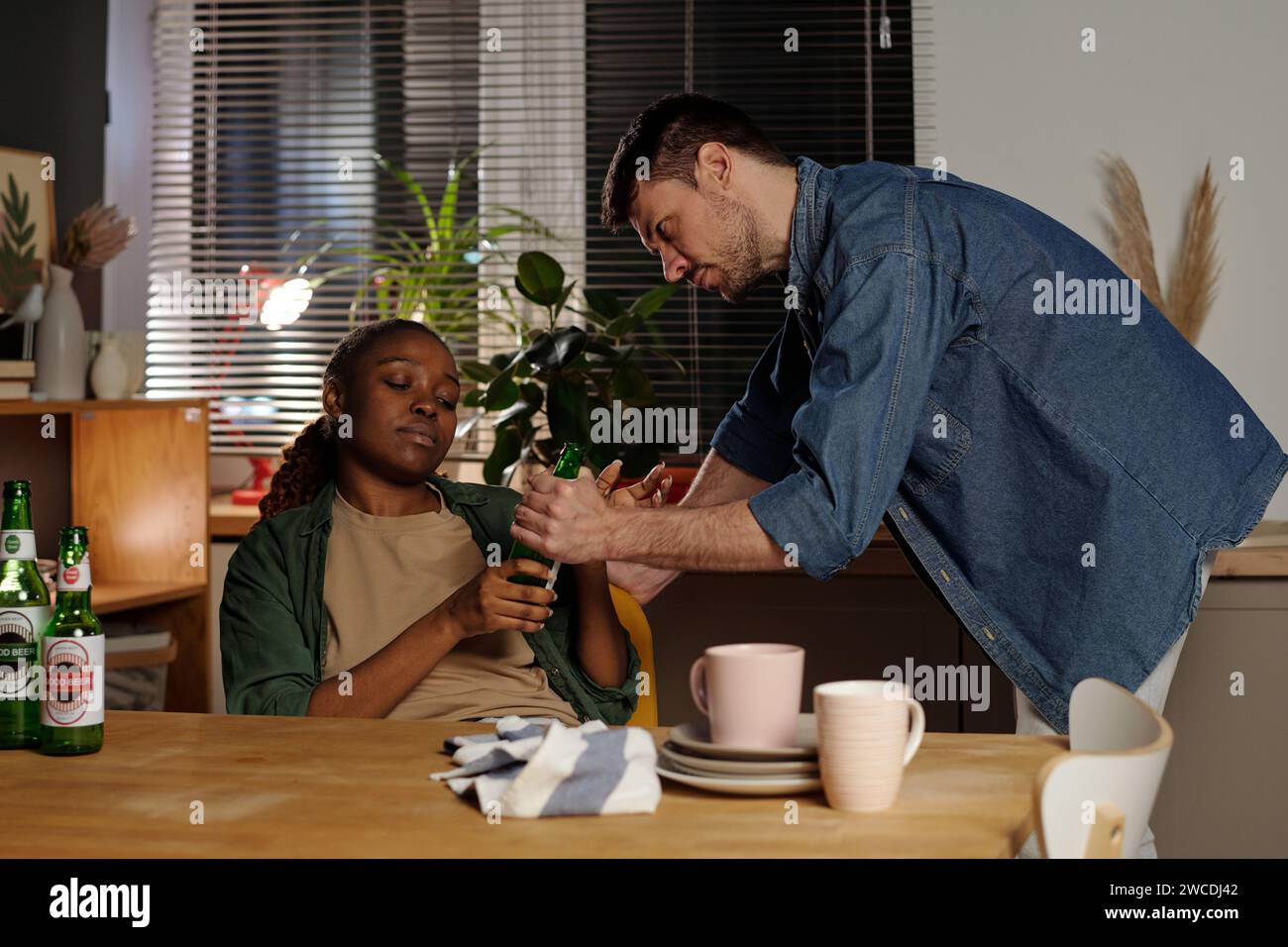 Young strict man taking bottle of beer from hands of his wife suffering ...