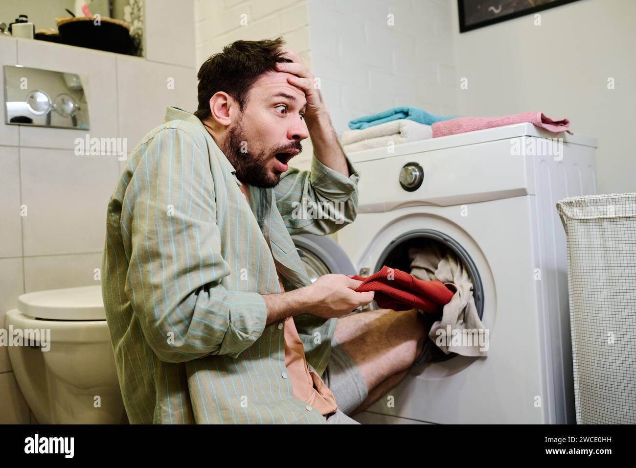Shocked young man pulling red piece of attire from washing machine and ...
