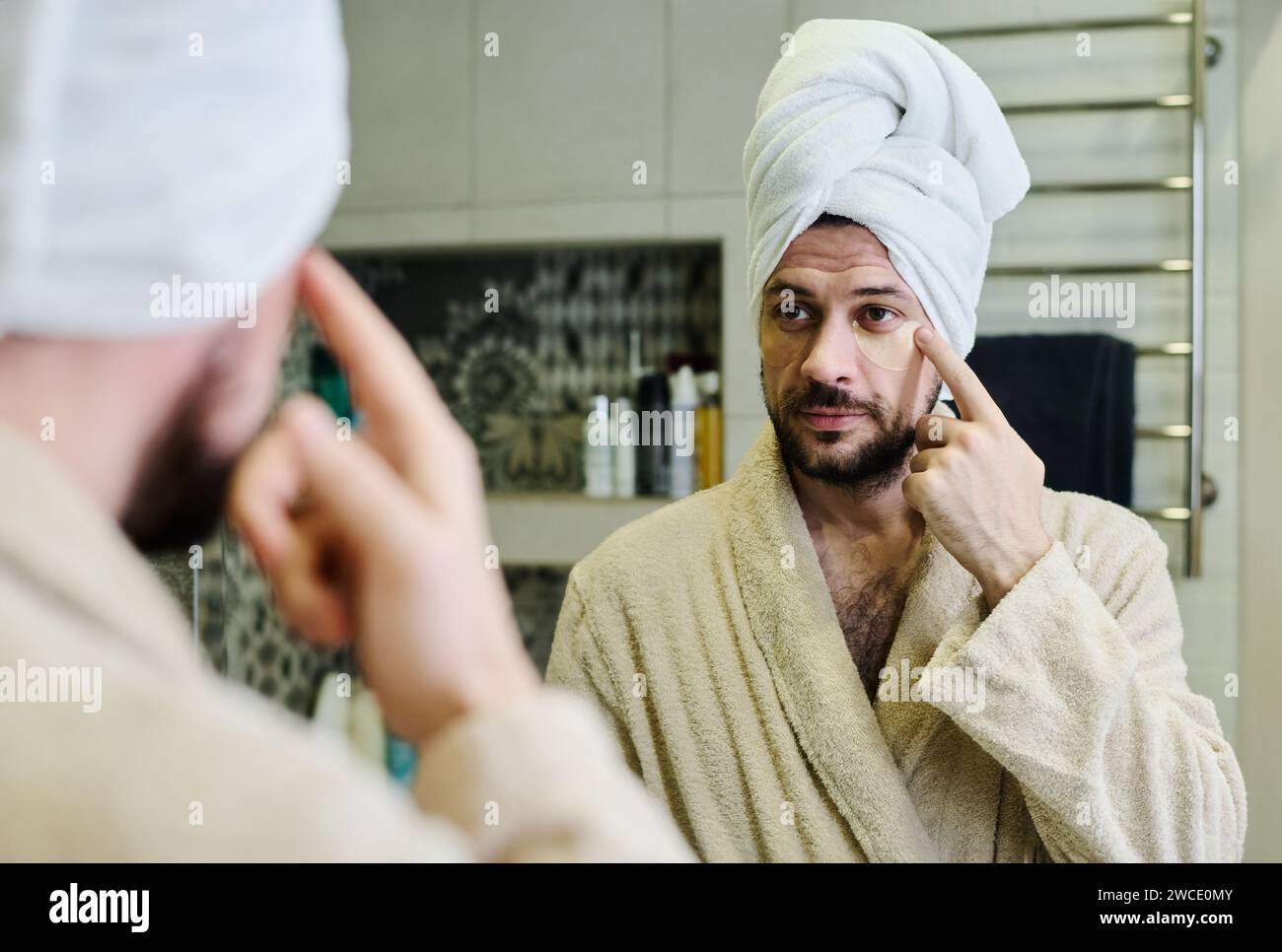 Young bearded man with towel on head putting moisturizing patches on ...