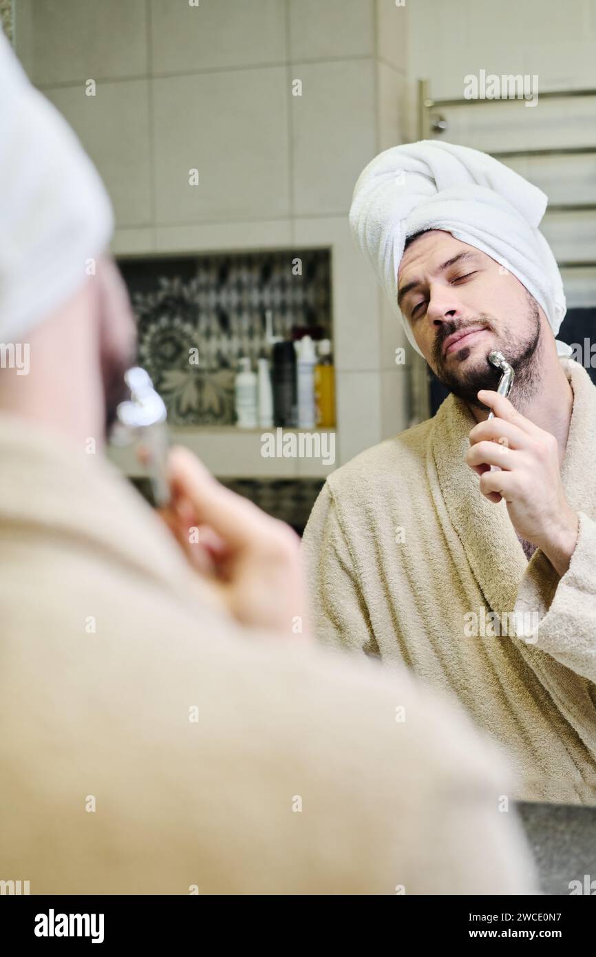 Young man in bathrobe taking pleasure in procedure of facial massage with Y-shaped ball roller ...