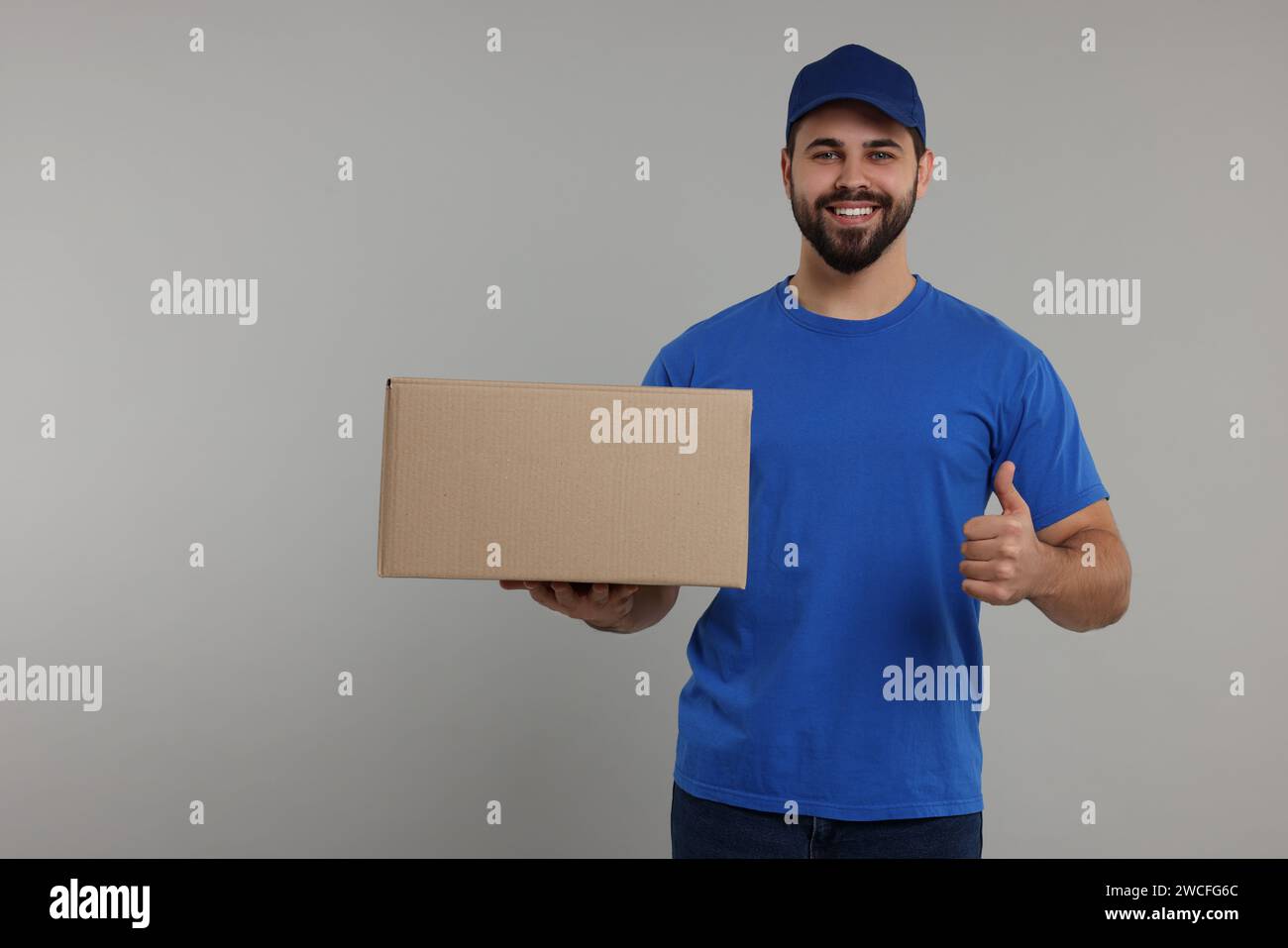 Happy courier with parcels showing thumb up on grey background, space ...