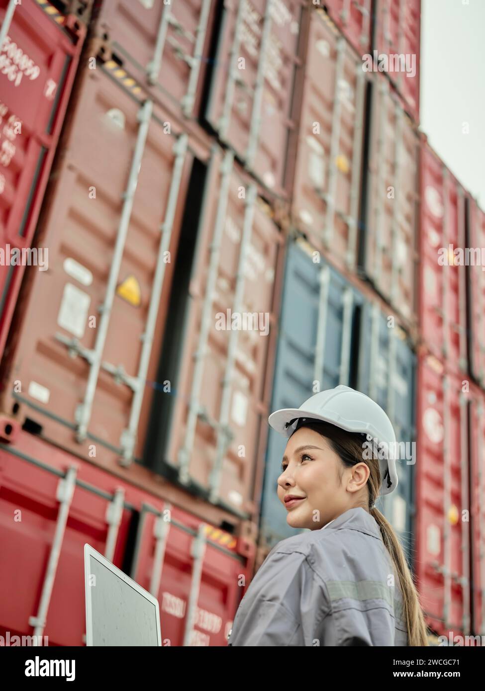 Portrait of asian woman worker holding Laptop and pointing with forefinger on shipping container ...