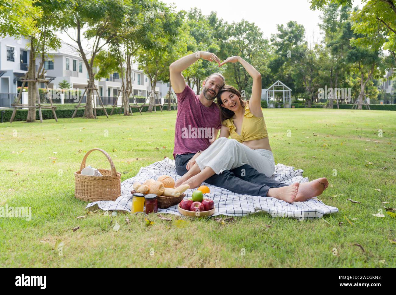 A couple seated on the grass, making a heart shape with their hands ...