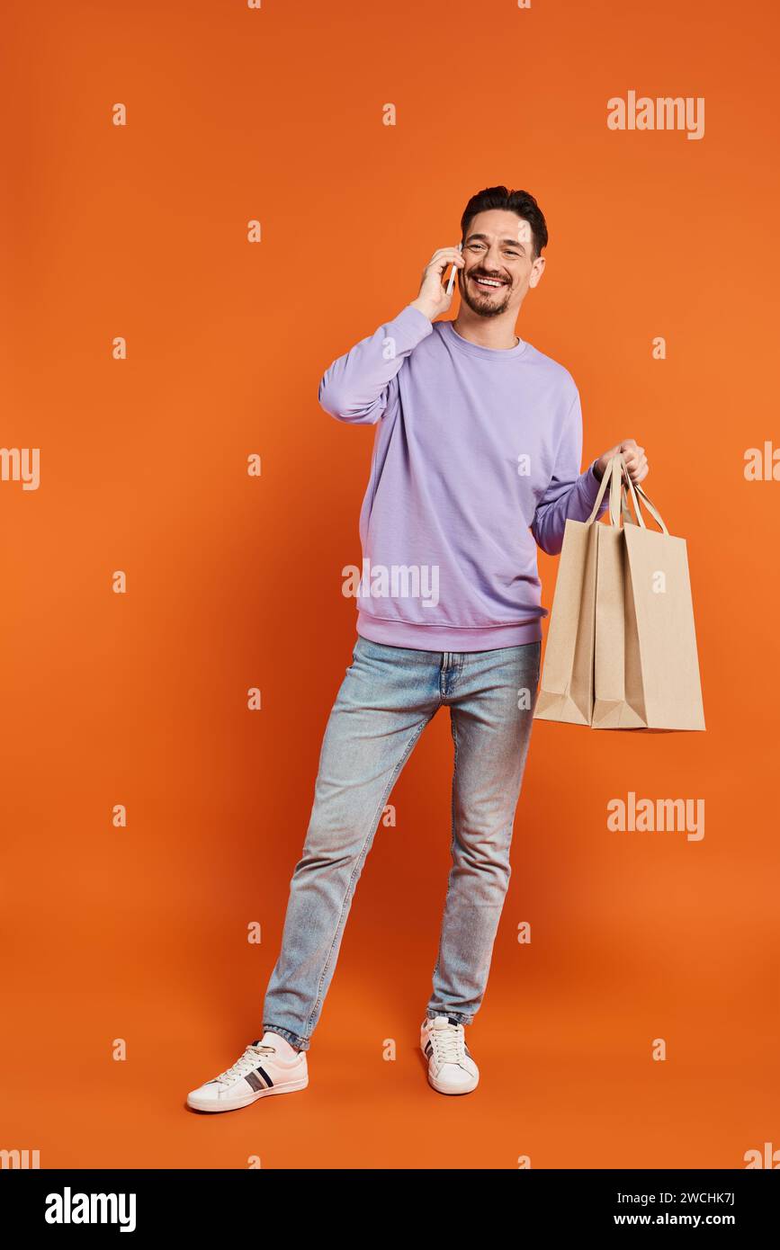cheerful man talking on the phone and carrying shopping bags on orange ...