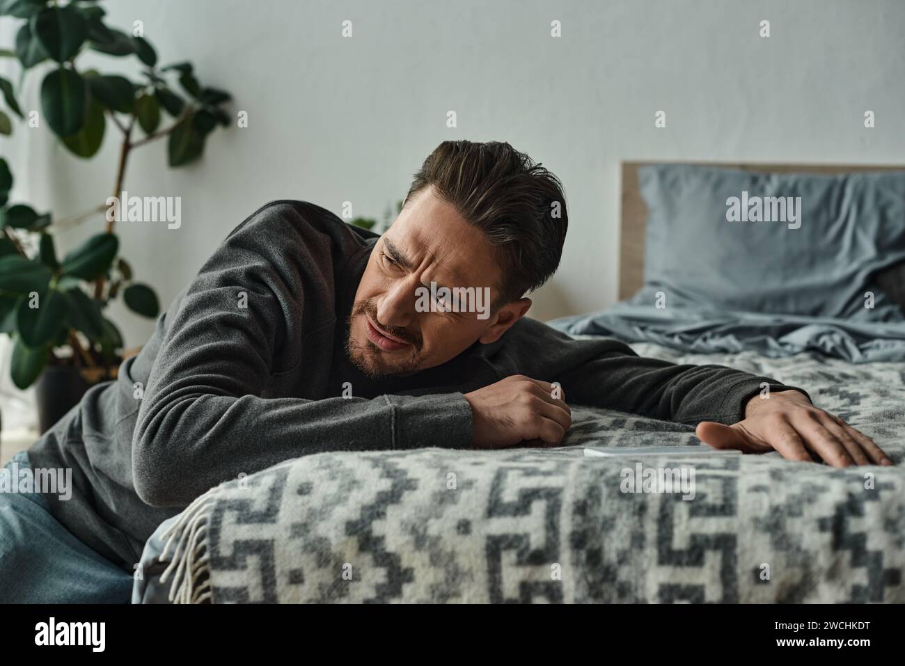 bearded man suffering from pain and leaning on grey blanket on bed in ...