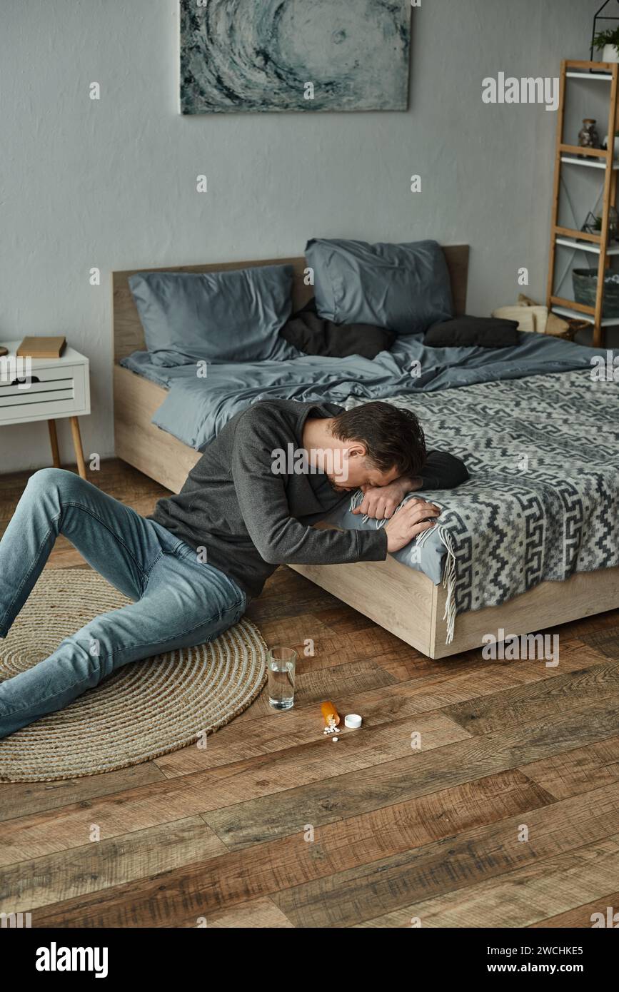 stressed bearded man sitting on floor near glass of water and pills in ...