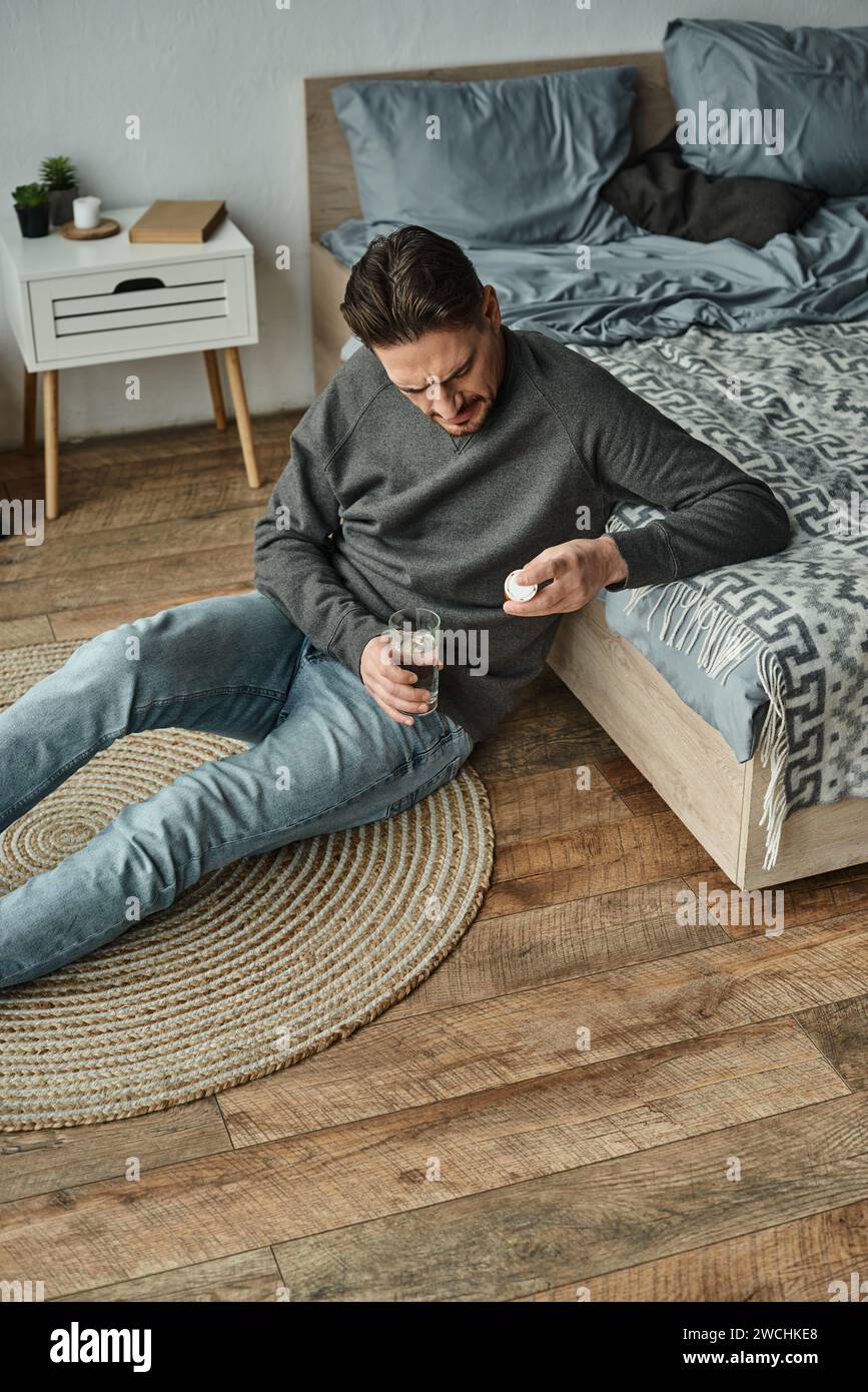 bearded man sitting on floor and holding glass of water and pills in ...