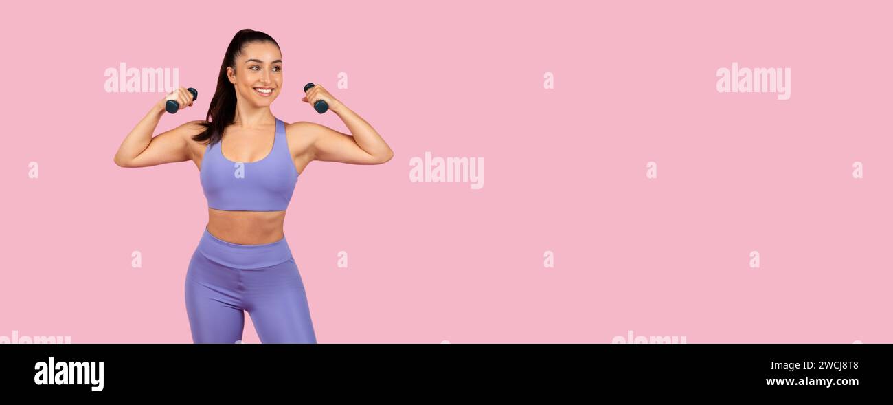 Happy young lady in sportswear doing exercise for hands with dumbbells ...