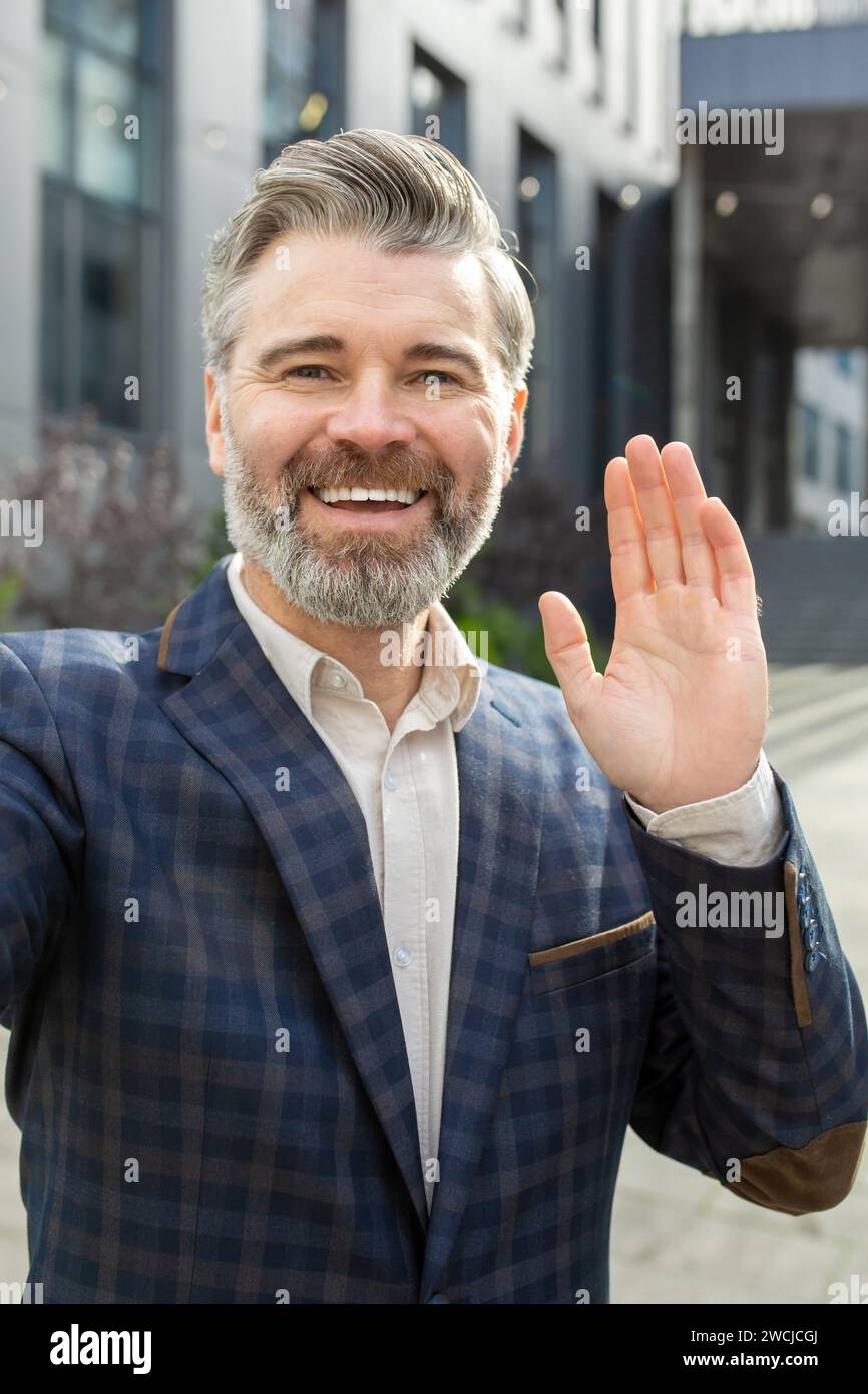 Smiling mature businessman in a stylish suit waving hello, standing ...