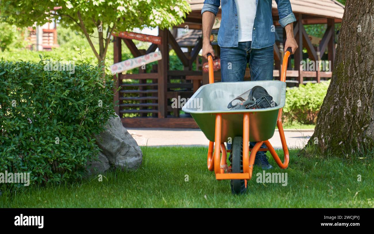 Unrecognizable male landscaper in jeans pushing wheelbarrow, while ...