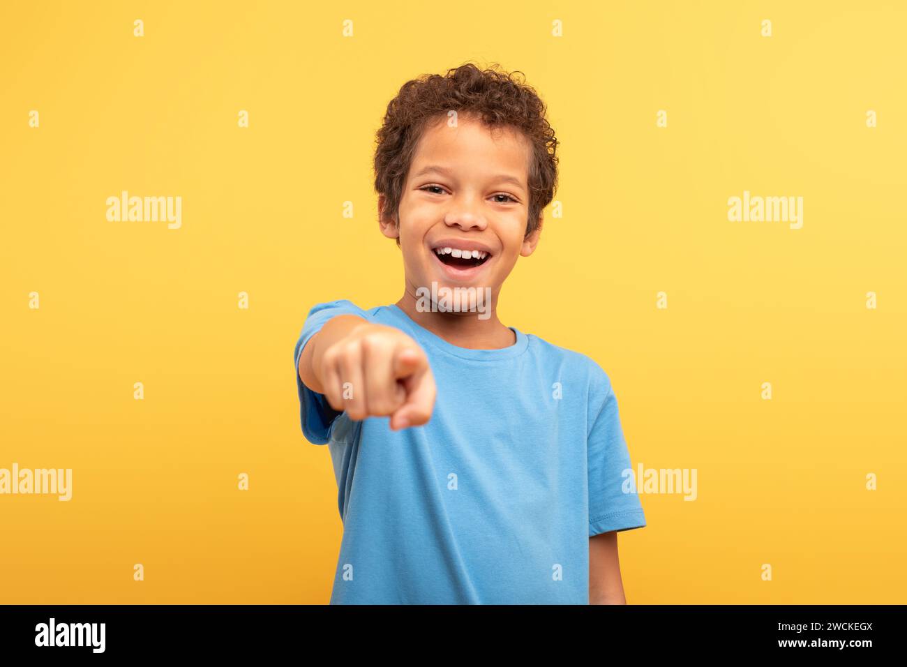 Laughing boy pointing to camera wearing blue t-shirt on yellow ...