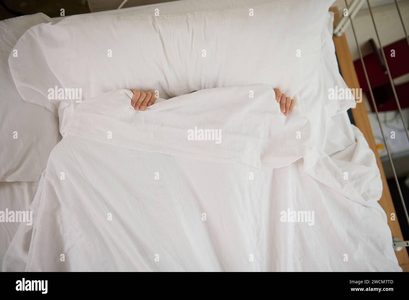 View from above of a scared little child hiding under white duvet while ...