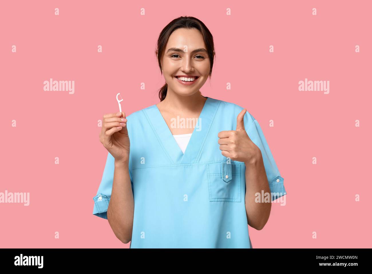 Young female dentist with floss toothpick showing thumb-up on pink ...