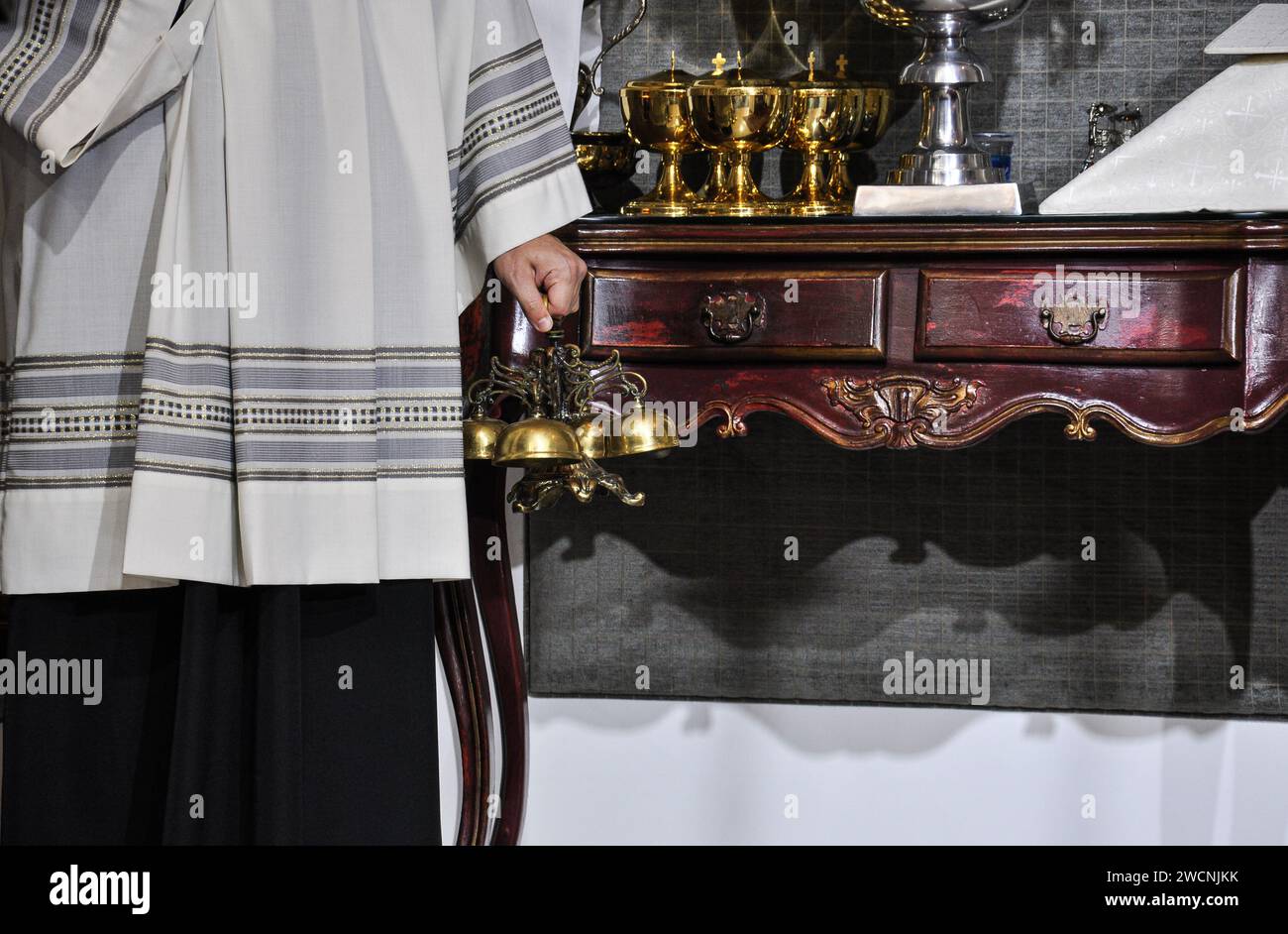 An acolyte, dressed in his white robe, holds a bell in his left hand ...
