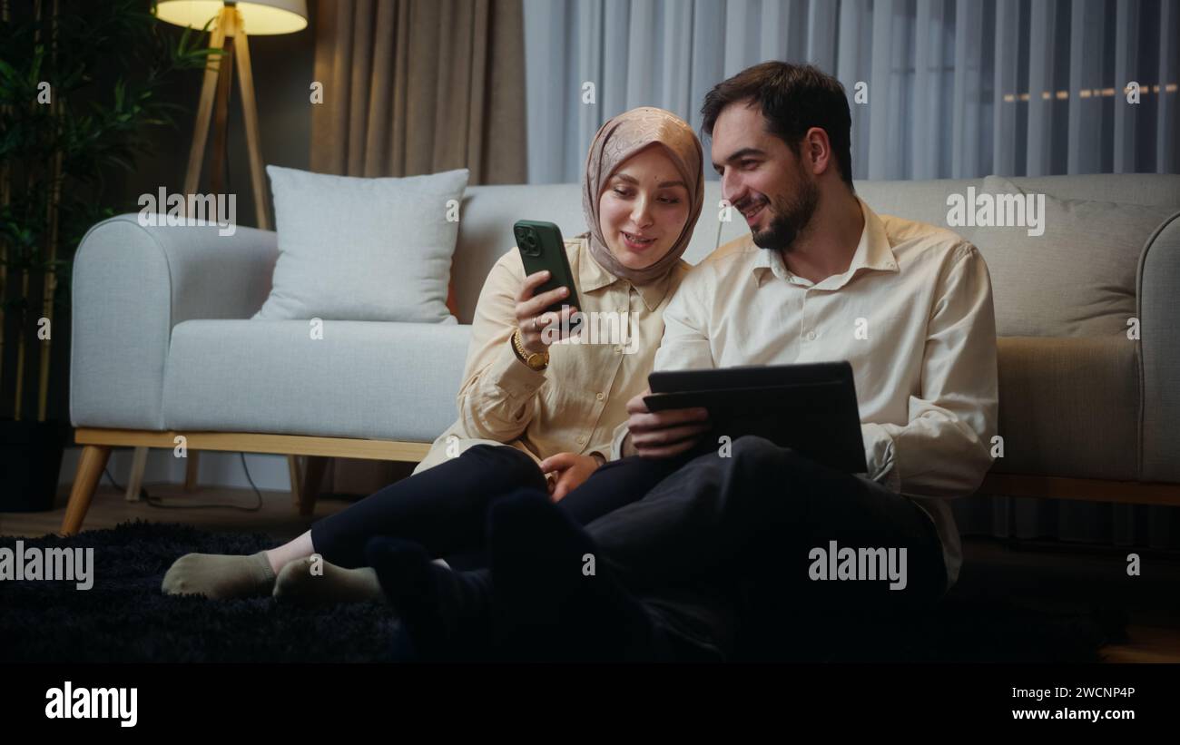 A married couple leaning against the sofa sitting on floor, woman using ...