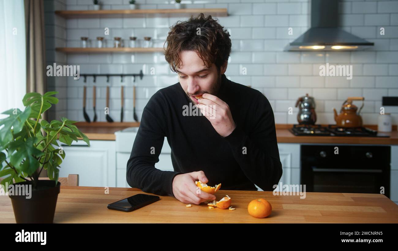 Young curly haired man sitting in the kitchen eating tangerines while ...