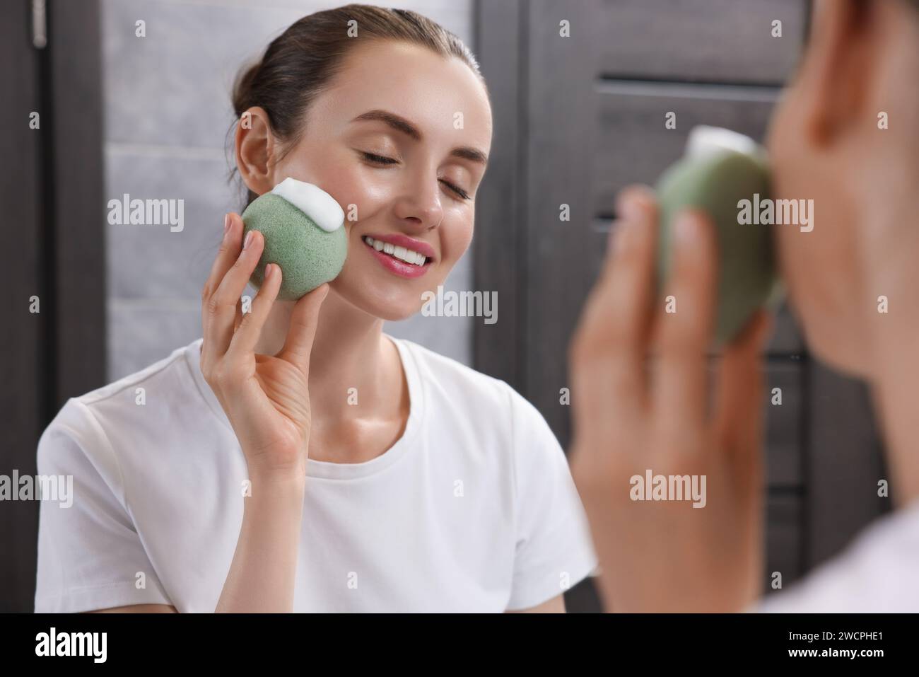 Happy young woman washing her face with sponge near mirror in bathroom ...