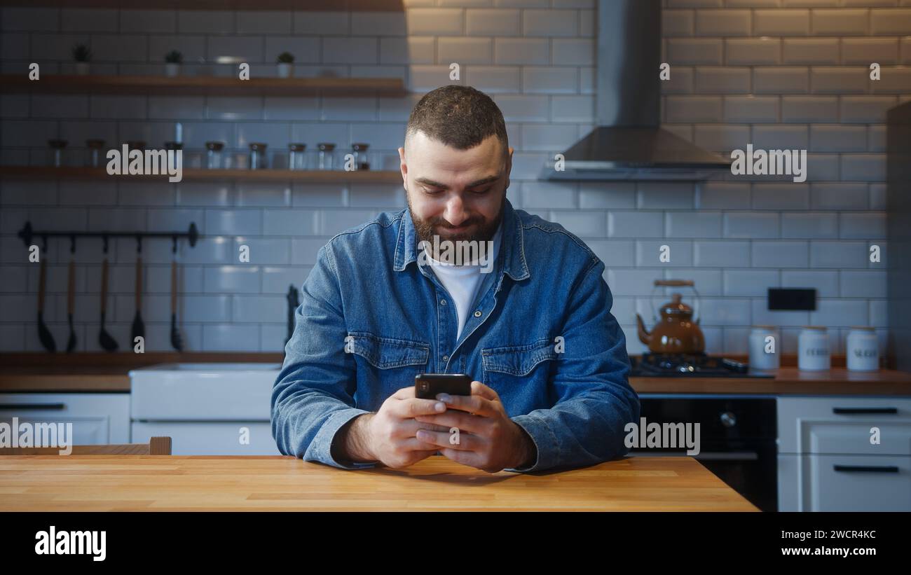 Bearded young adult man sitting against the kitchen counter typing text ...