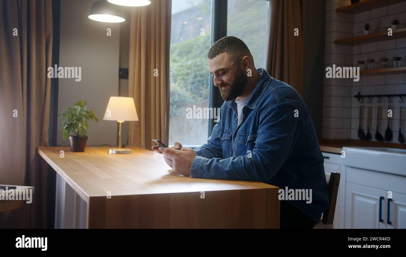 Bearded young man sitting against the kitchen counter typing text ...