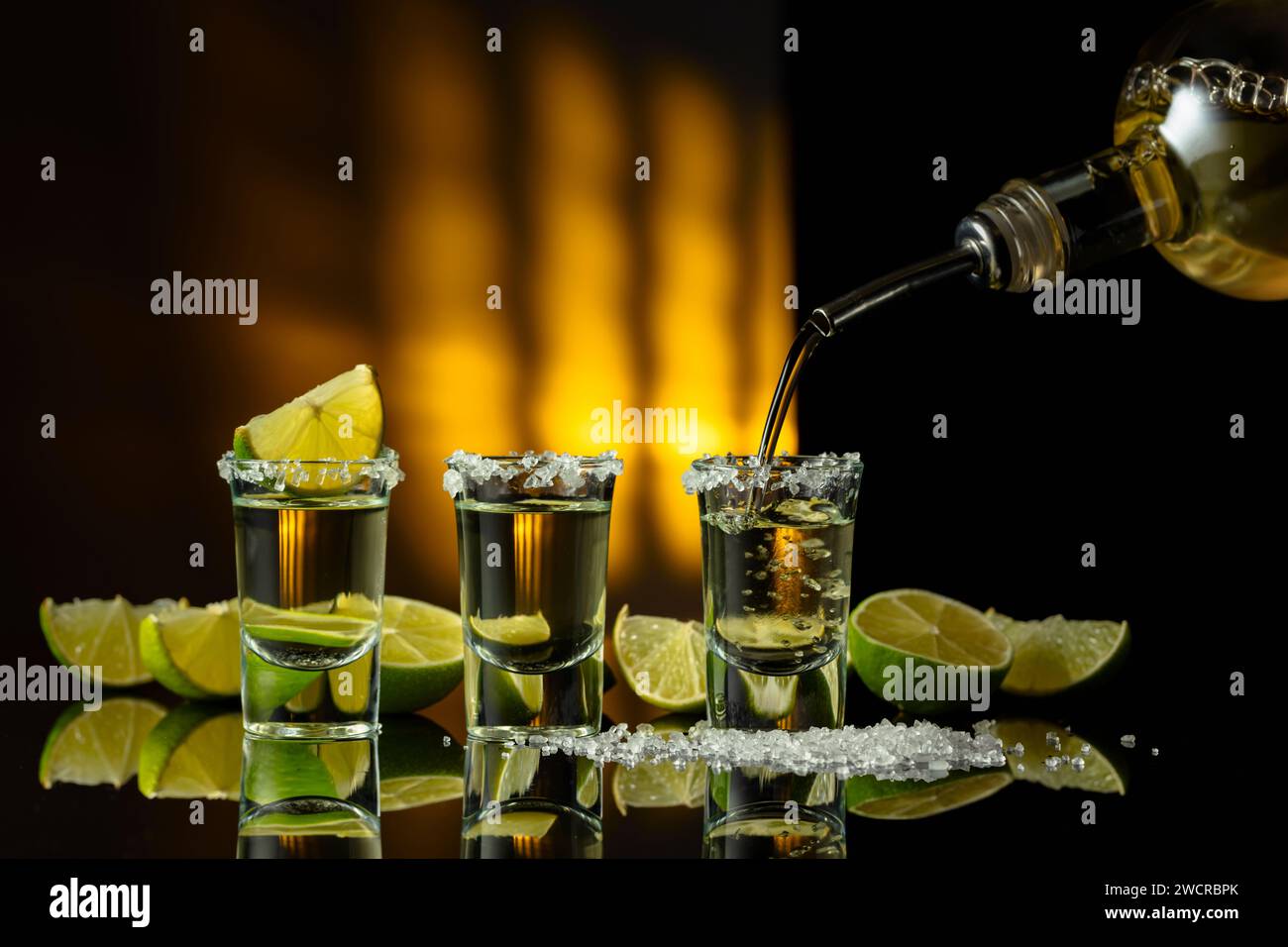 Gold tequila with sea salt and lime slices on a black reflective ...