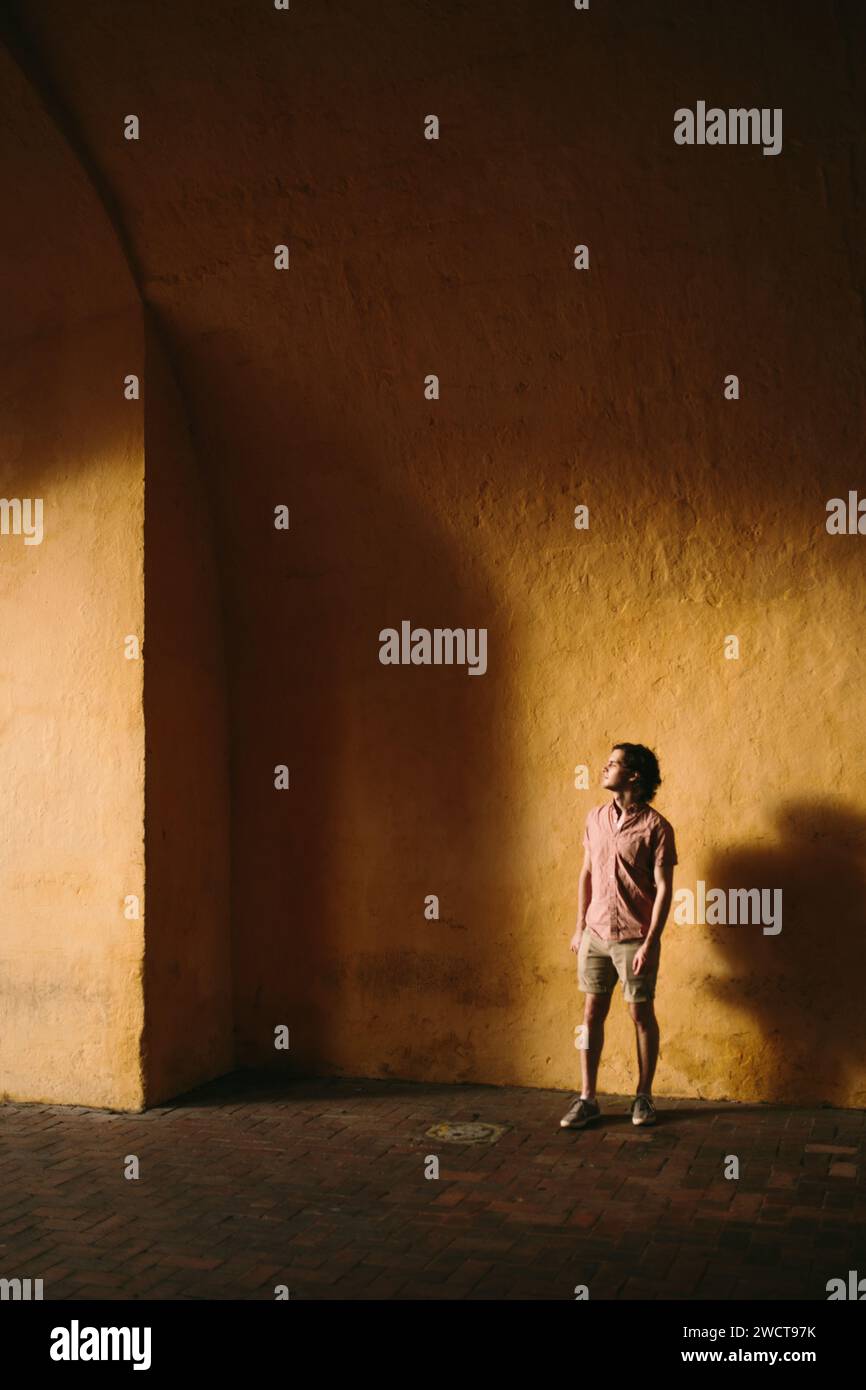 A contemplative young man stands alone against a warm-toned wall bathed ...
