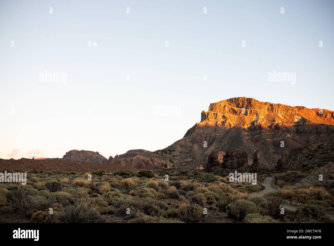 The setting sun casts a vibrant orange glow on a rugged desert mountain ...
