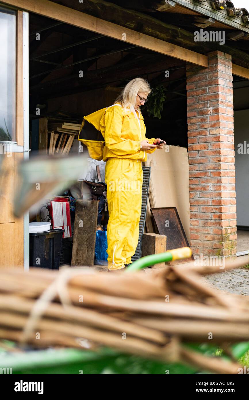 Focused female Beekeeper in yellow overall using mobile phone while ...