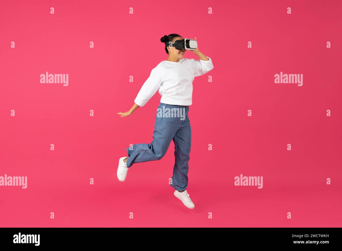 Excited young asian woman wearing VR headset jumping on pink background ...