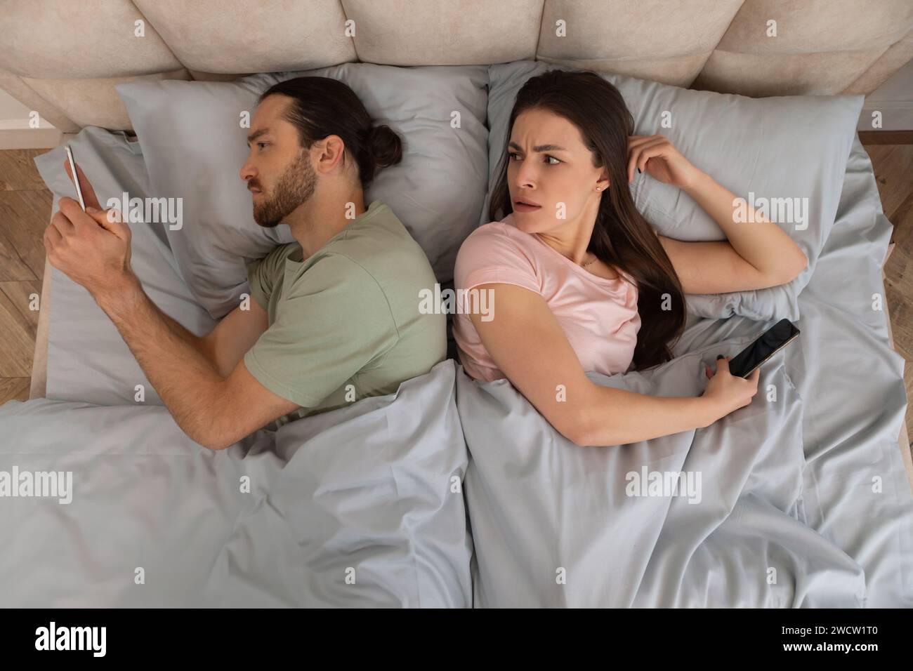 Sad young woman turning to husband chatting on his smartphone in bed at ...