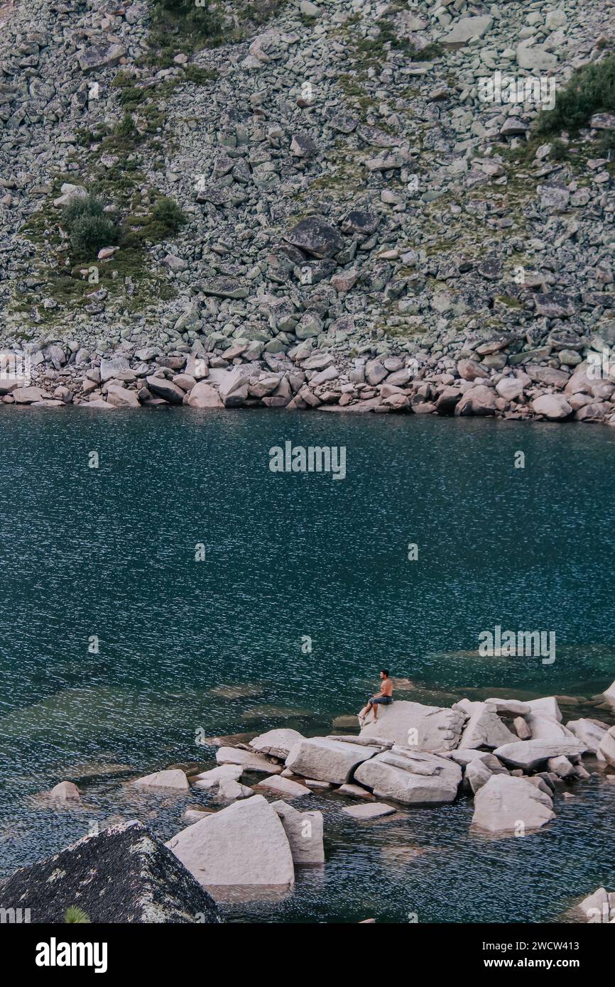 Man sits on large stone protruding from water near shore of emerald ...