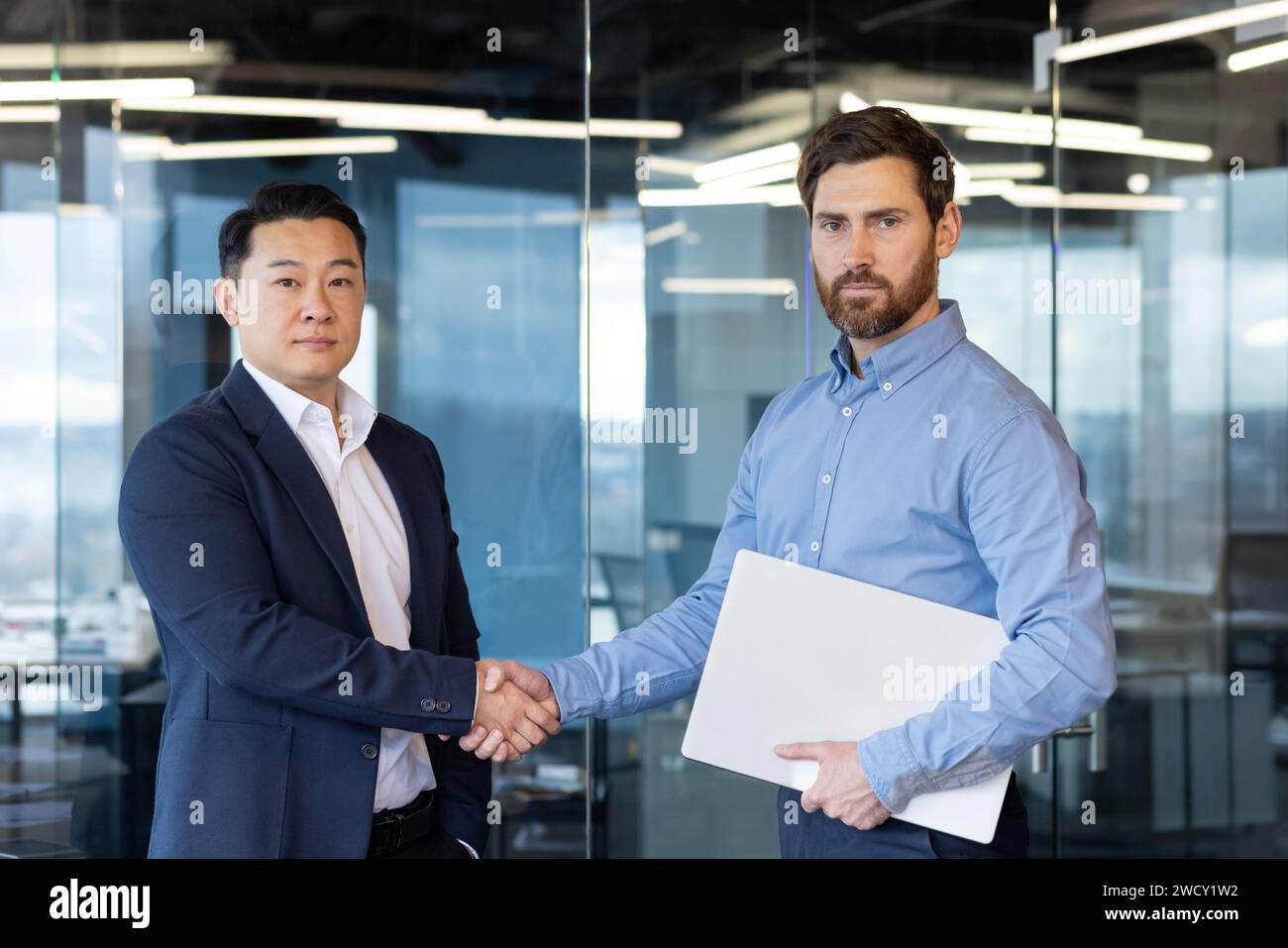 Two diverse businessmen shaking hands in a bright office setting, illustrating partnership and ...