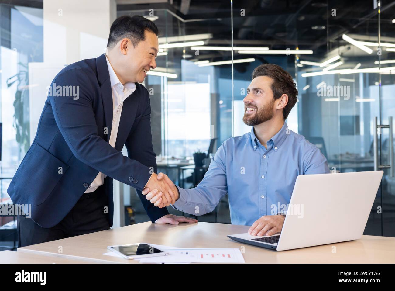 Two professional businessmen in a modern office setting engaging in a friendly handshake ...