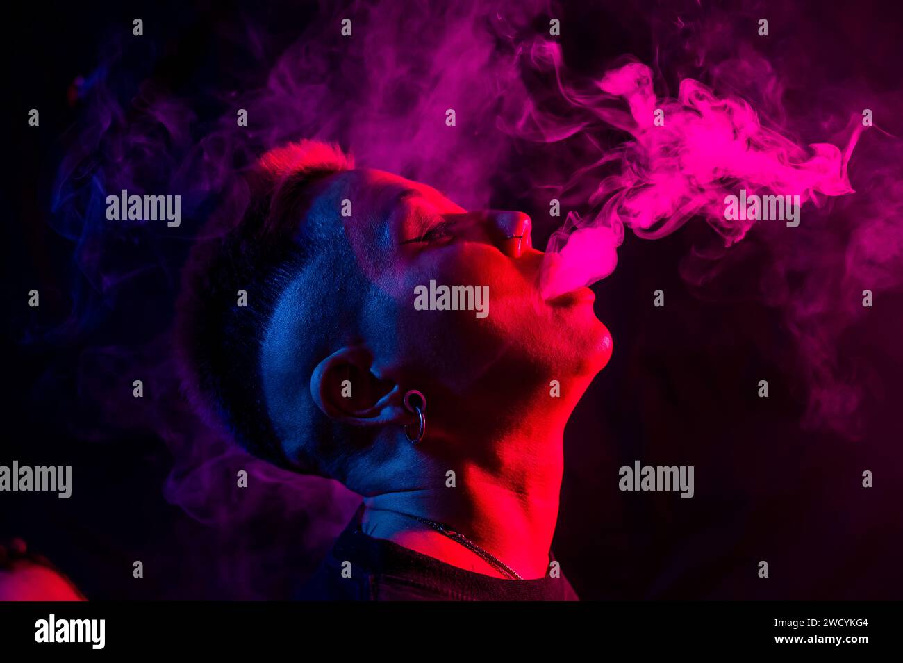 Portrait asian woman with shaved temples smokes vape in neon light ...