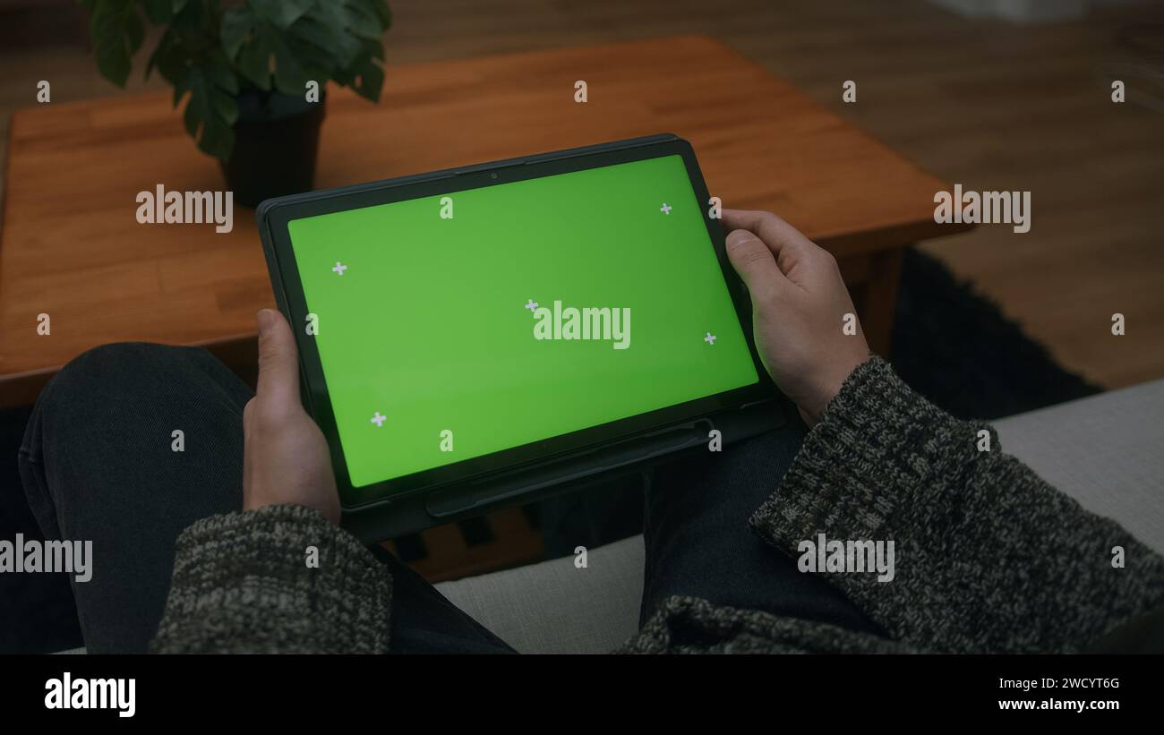 Young man scrolling and tapping center on tablet with green screen mock up display. Male sitting ...