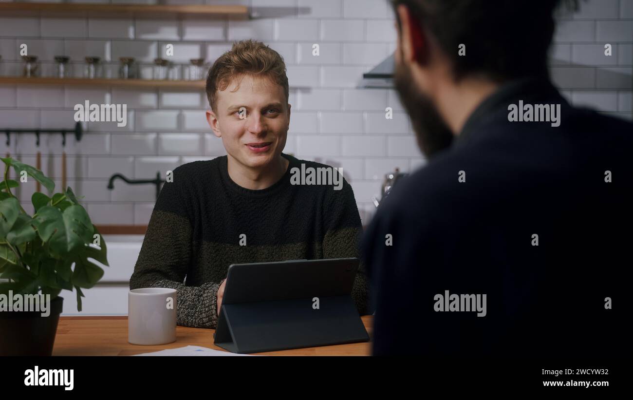 Two happy co-worker men are sitting in the kitchen having a fun chat ...