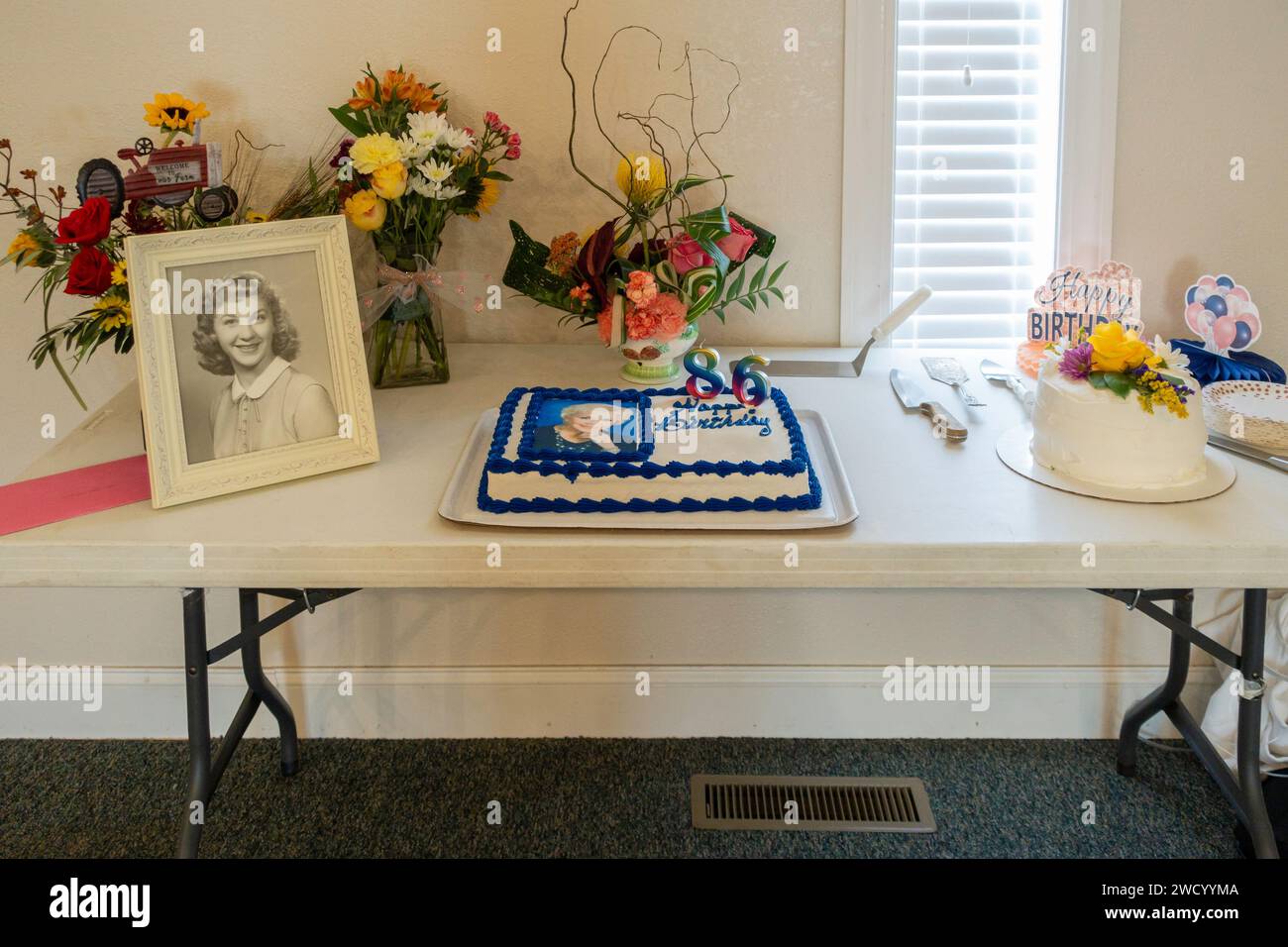 A serving table with two birthday cakes, a photograph, vases of flowers ...