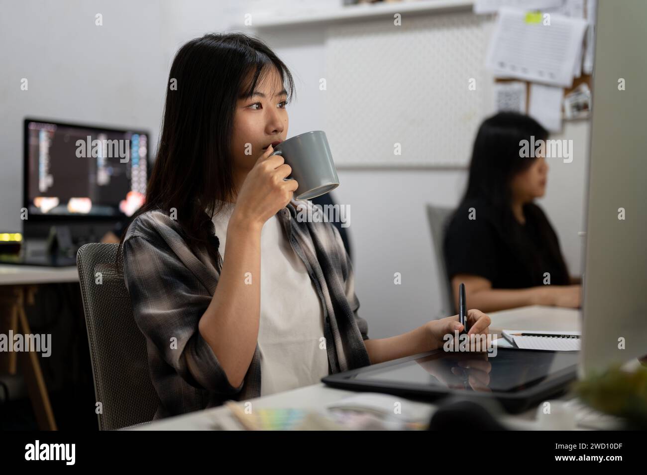Concentrated young Asian female graphic designer is sipping coffee ...